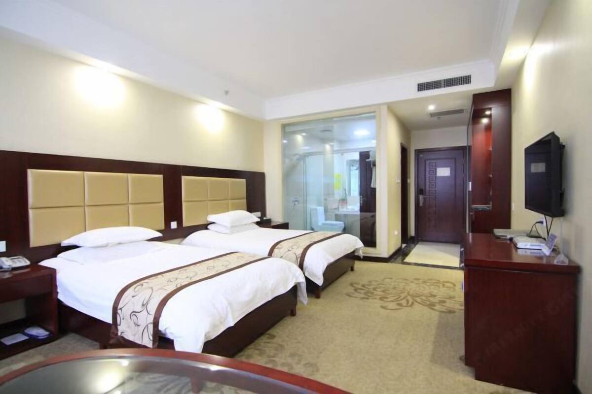 Quarto de hotel com duas camas, uma escrivaninha, televisão e um banheiro visível por uma porta de vidro.