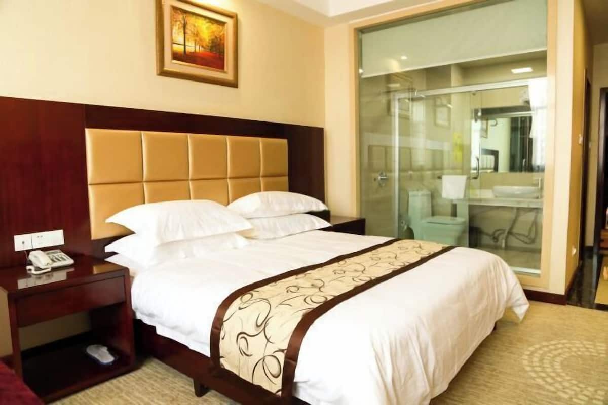 Quarto de hotel com uma cama grande, mesa de cabeceira com telefone, box de chuveiro de vidro e um quadro na parede.