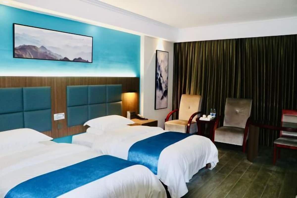 Quarto de hotel com duas camas, uma escrivaninha, uma cadeira e uma janela grande com cortinas.