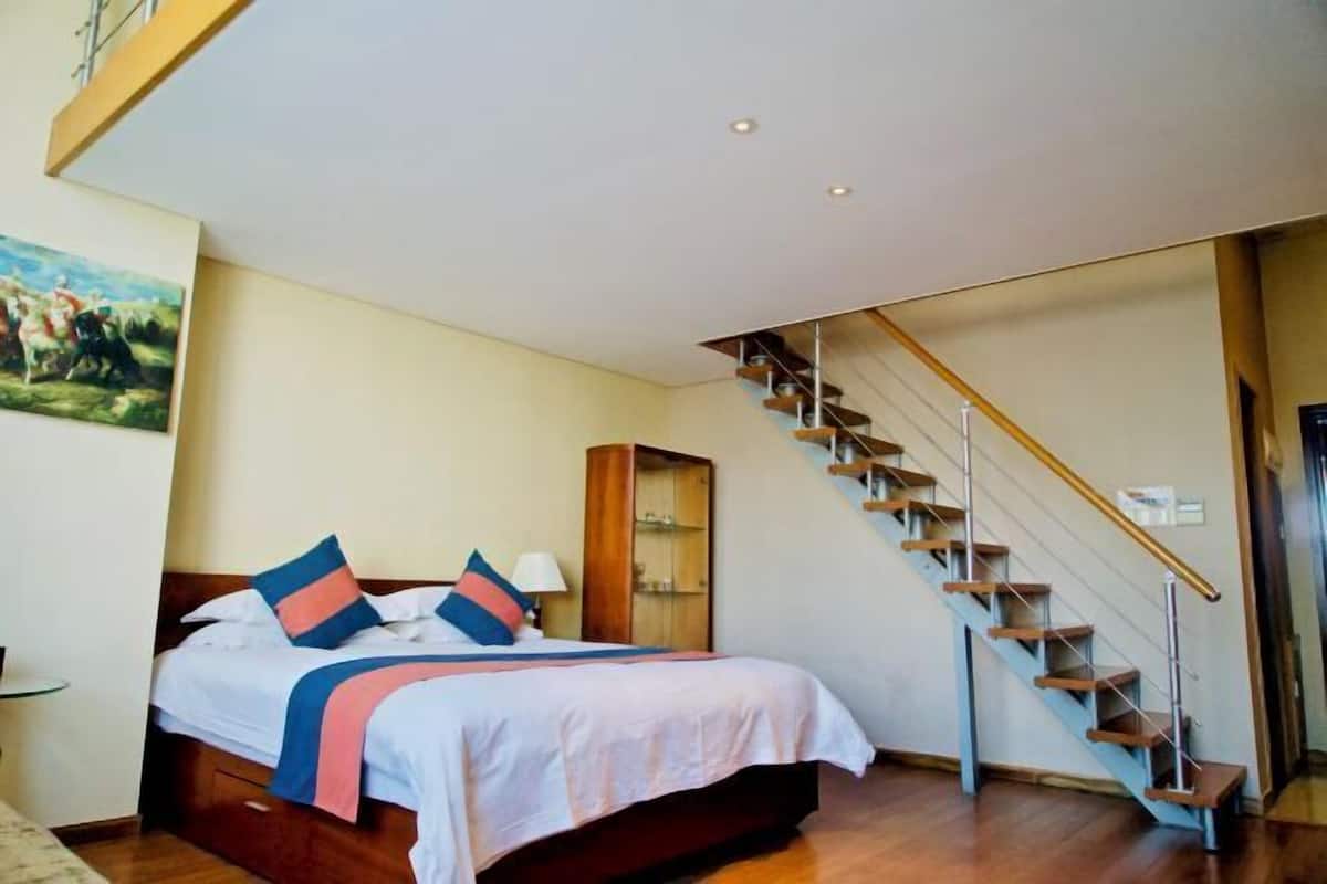 Um quarto com uma cama, uma mesa de cabeceira, uma luminária, um guarda-roupa de madeira e uma escada.