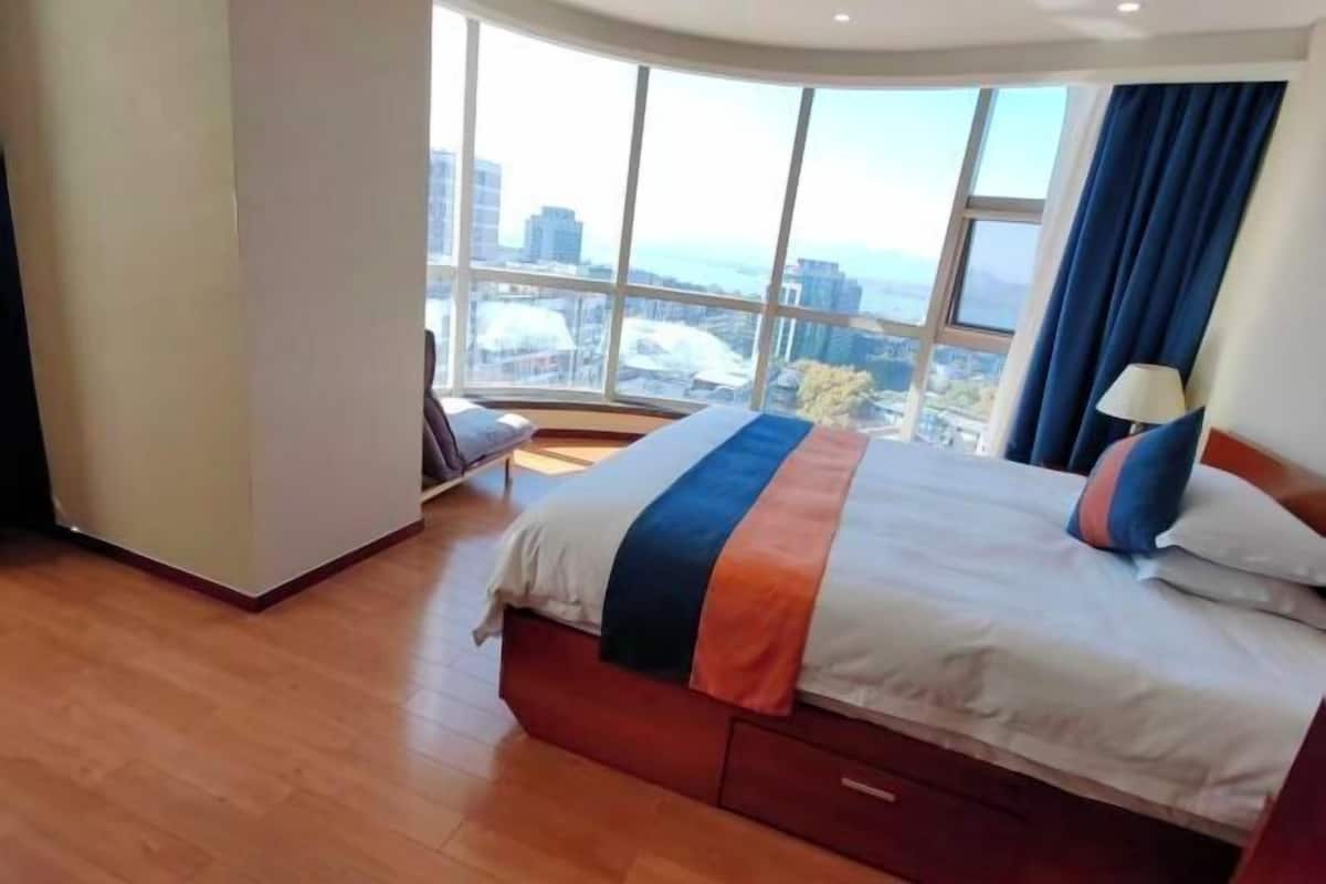 Quarto de hotel com uma janela grande, uma cama com manta listrada, mesa de cabeceira e abajur.
