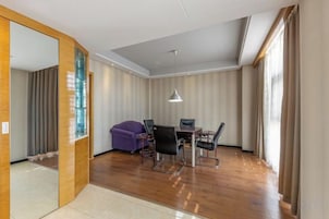 Quarto de hotel com piso de madeira, área de jantar com mesa e cadeiras, sofá roxo e uma janela grande com cortinas.