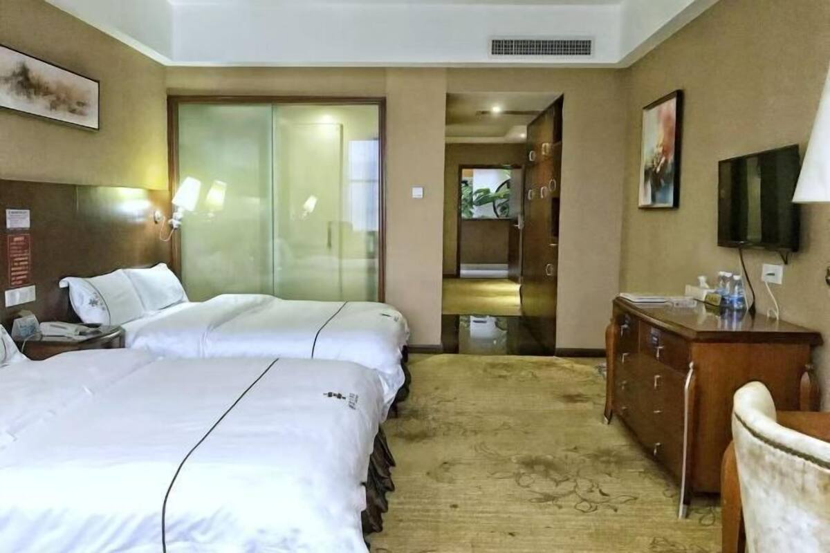 Quarto de hotel com duas camas, uma escrivaninha, uma televisão e uma porta deslizante que dá para outro cômodo.