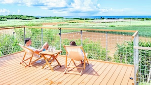 Um deck de madeira com duas cadeiras dobráveis e uma mesa, com vista para uma paisagem verde e um corpo d'água.