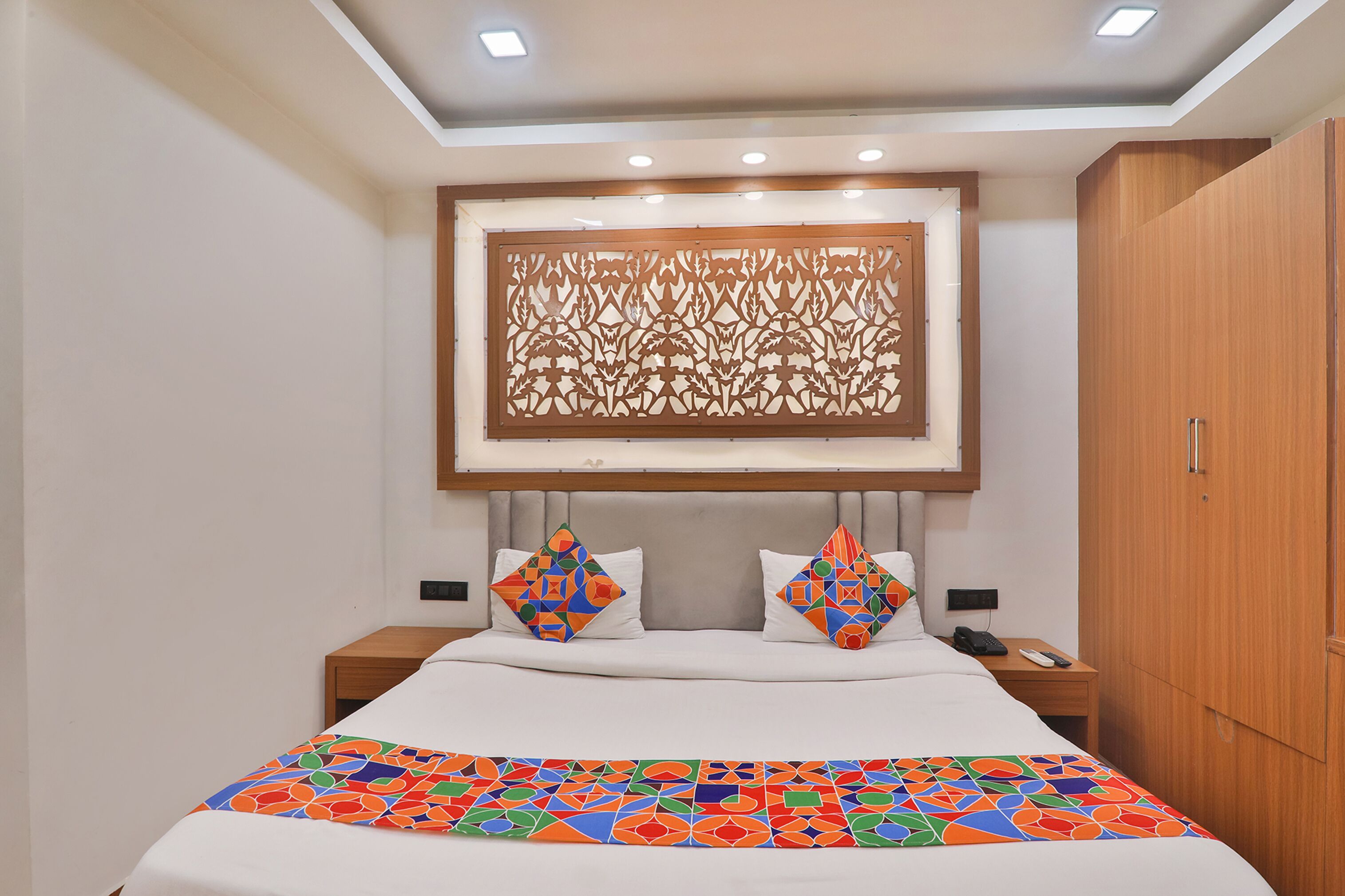 Quarto de hotel moderno com uma cama grande, cabeceira decorativa, guarda-roupa de madeira e mesa de cabeceira com telefone e controle remoto.