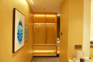 Corredor de hotel com armário de madeira, pintura abstrata emoldurada e banheiro com pia e porta-toalhas.
