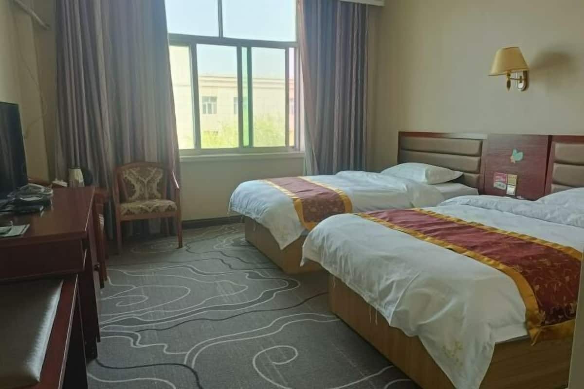 Quarto de hotel com duas camas, uma escrivaninha, uma cadeira e uma janela com cortinas.