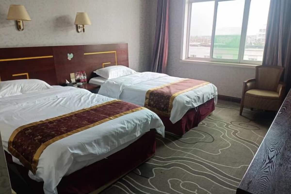 Quarto de hotel com duas camas, cabeceira, luminárias fixadas na parede, uma cadeira e vista para o exterior.