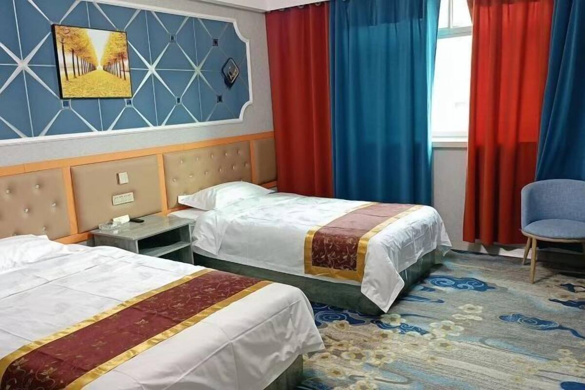Quarto de hotel com duas camas, uma mesa de cabeceira, uma cadeira e uma janela grande com cortinas vermelhas.