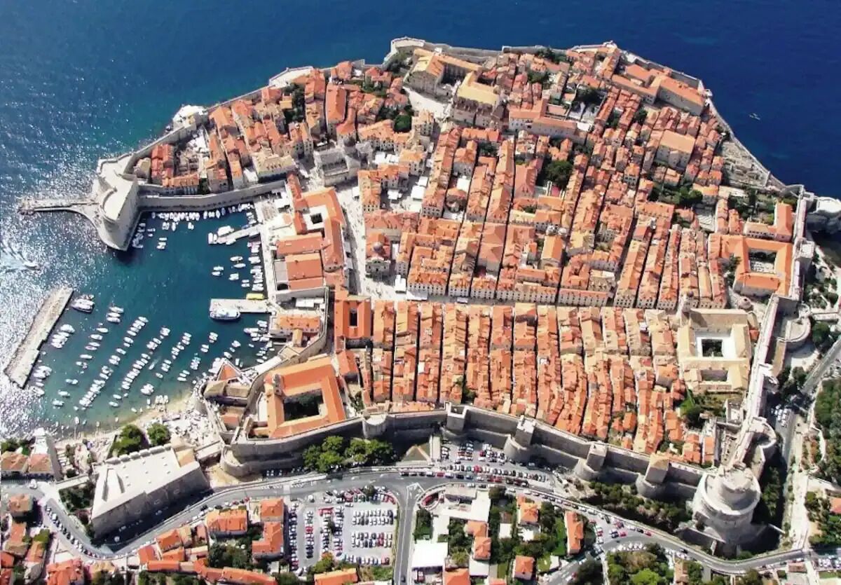 Vista aérea de uma vila costeira com uma muralha fortificada, um porto com barcos e um conjunto denso de edifícios com telhados vermelhos.