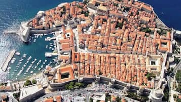 Vista aérea de uma vila costeira com uma muralha fortificada, um porto com barcos e um conjunto denso de edifícios com telhados vermelhos.