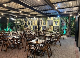Restaurante ao ar livre com piso de tijolos, mesas e cadeiras de madeira, e área coberta com telhado de metal.