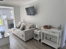 Uma sala de estar moderna com um sofá cinzento, uma mesinha de apoio branca, uma TV fixada na parede e uma pequena escrivaninha branca com gavetas.
