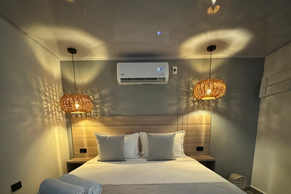 Quarto com uma cama grande, duas luminárias de cabeceira, um aparelho de ar condicionado e um ponto de luz fixado na parede.