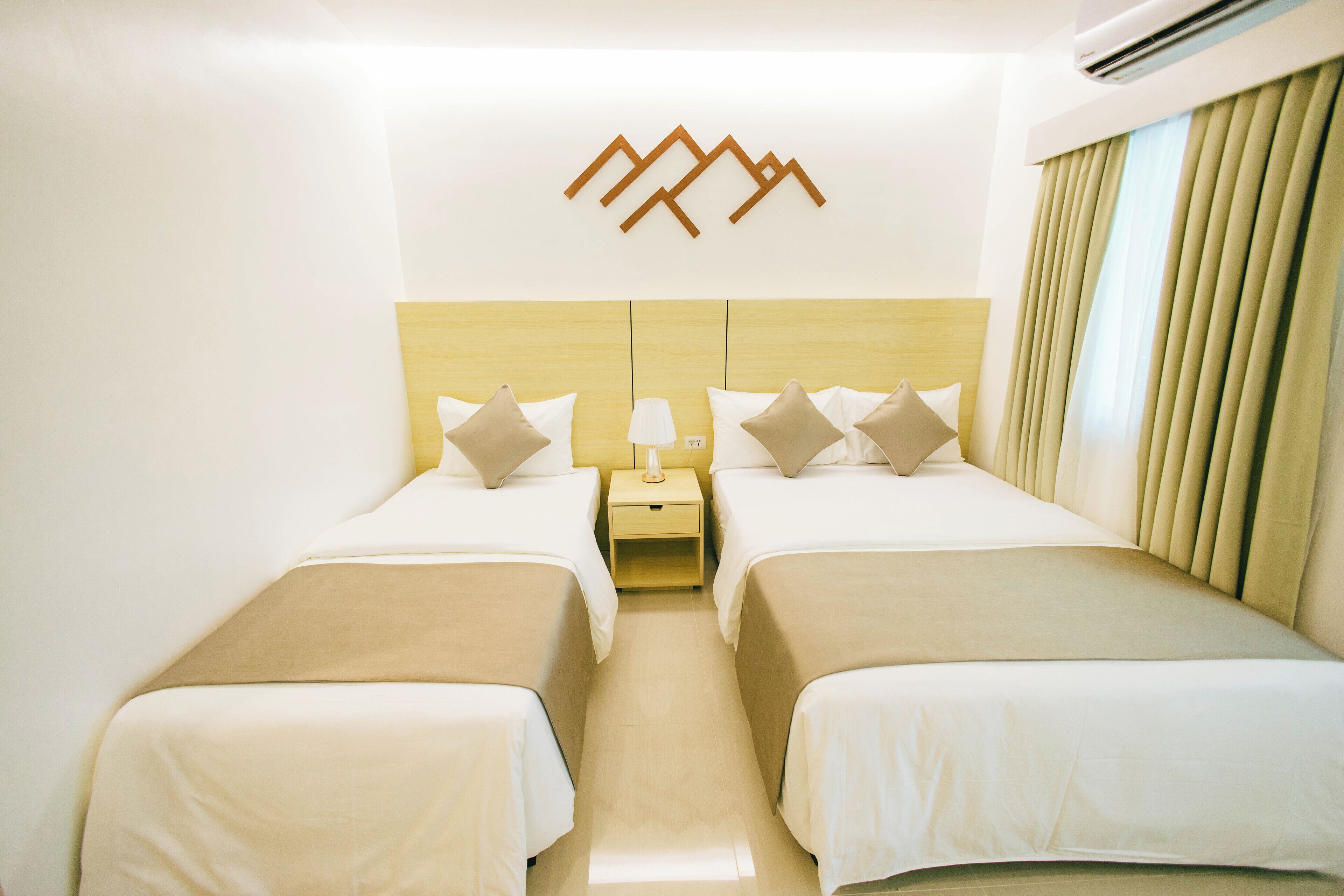 Quarto de hotel com duas camas, cabeceira de madeira, ar-condicionado fixado na parede e mesa de cabeceira com abajur.