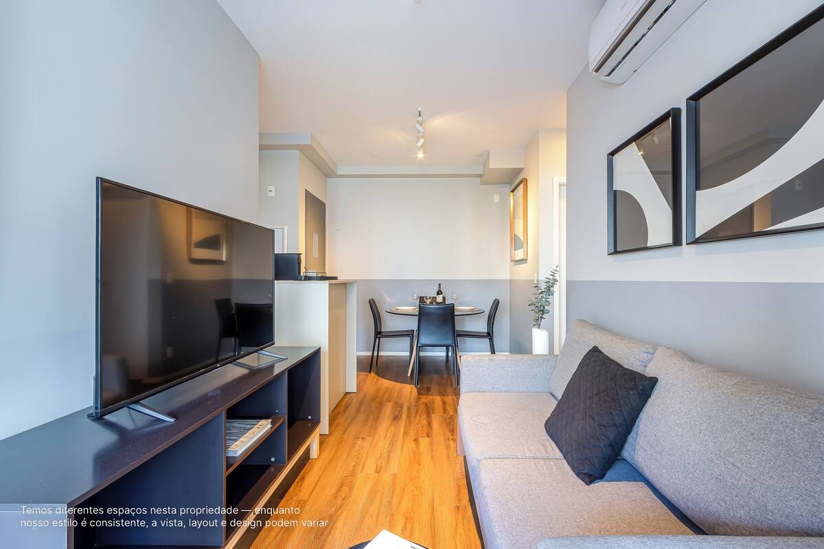 Uma sala de estar moderna com televisão de tela plana, um sofá cinza e uma área de jantar com mesa e cadeiras.
