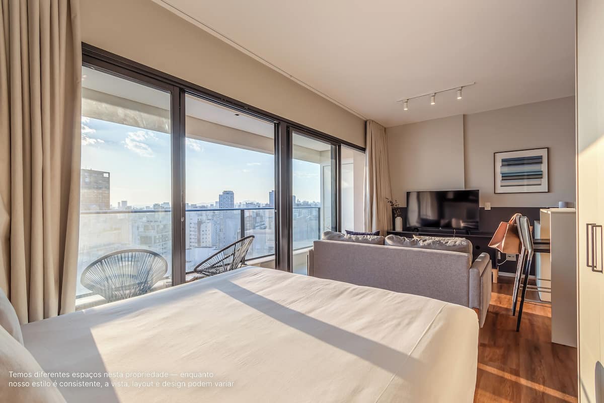 Quarto de hotel moderno com uma cama grande, área de estar, televisão e vista para a cidade.