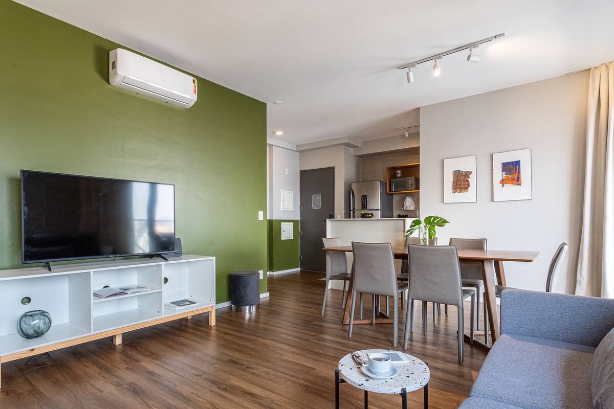 Uma sala de estar moderna com televisão de tela plana, uma área de jantar com mesa e cadeiras, e uma cozinha ao fundo.