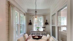 Uma sala de jantar com uma mesa de madeira, cadeiras brancas, um candelabro e uma janela com cortinas.