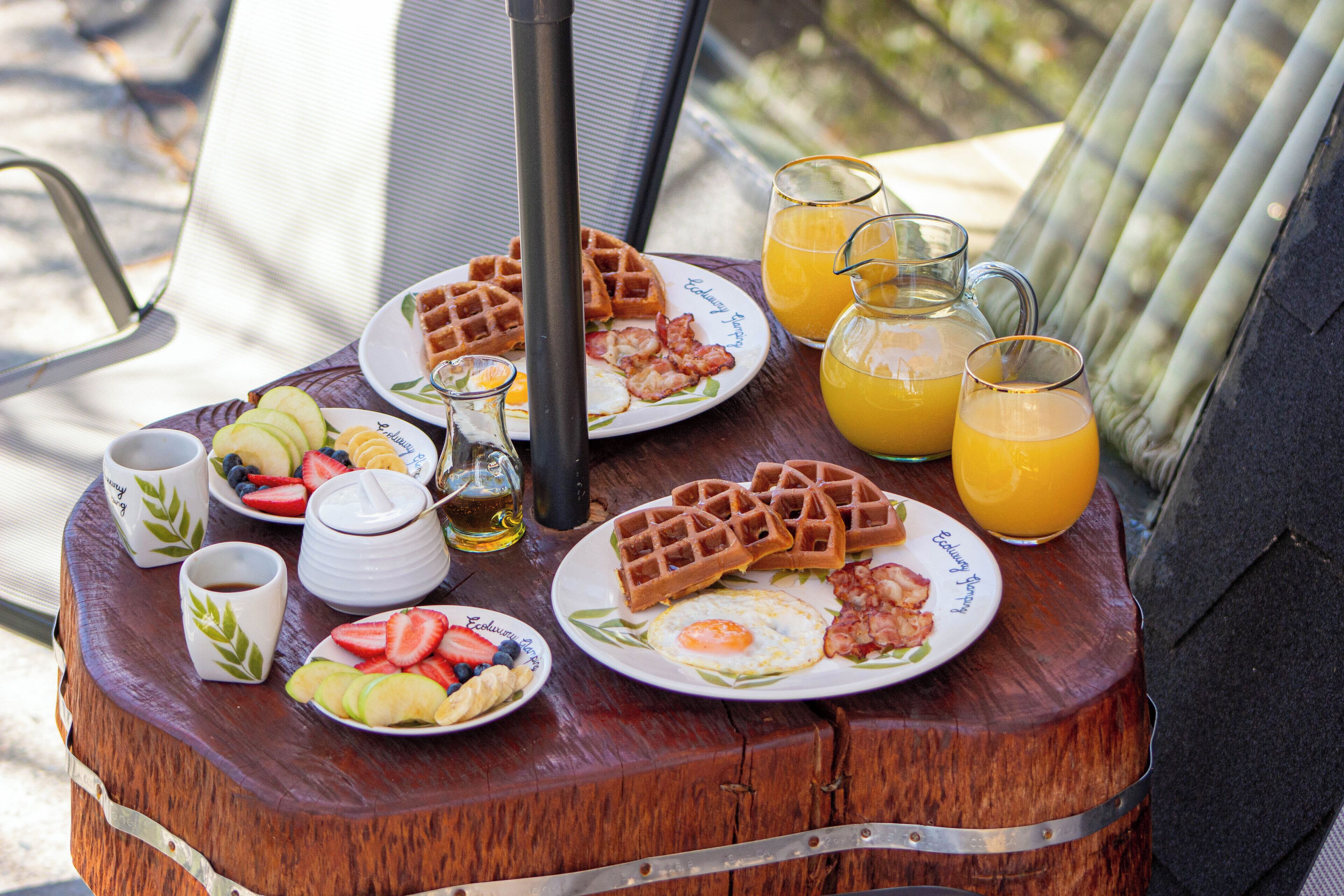 Uma mesa de café da manhã com waffles, ovos, bacon e frutas. Também há dois copos de suco de laranja e uma xícara de café.