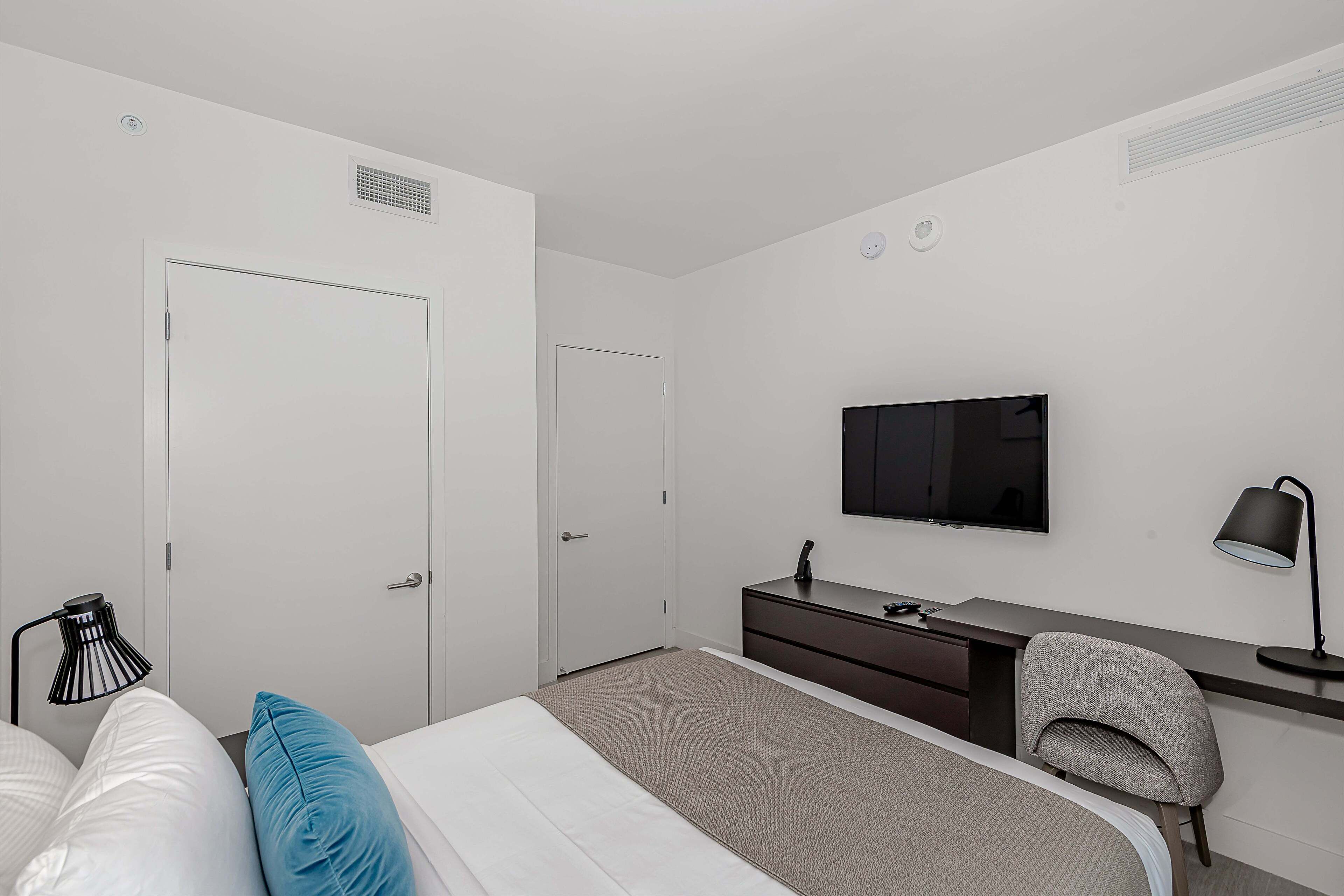 Quarto de hotel moderno com cama, escrivaninha, cadeira, televisão e abajur.