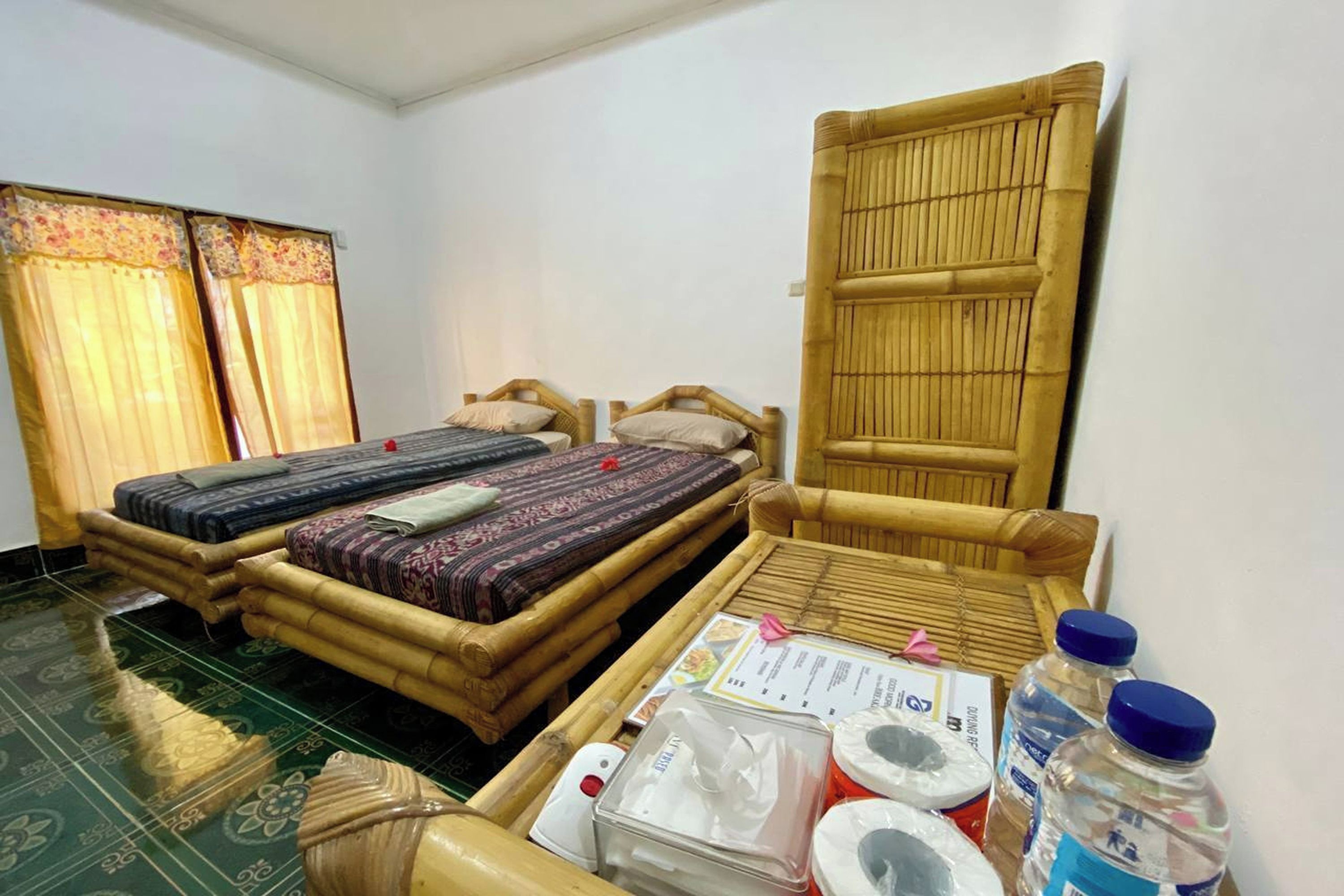 Quarto com duas camas de madeira, um separador de bambu, piso com padrão verde e uma mesa com cardápio e garrafas de água.