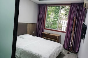 Uma cama de solteiro com cabeceira de madeira, uma pequena prateleira de madeira, uma janela com cortinas roxas e um cabideiro de madeira.