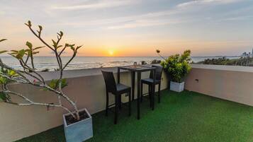 Terraço com vista para o pôr do sol sobre o oceano, duas cadeiras e plantas em vasos.