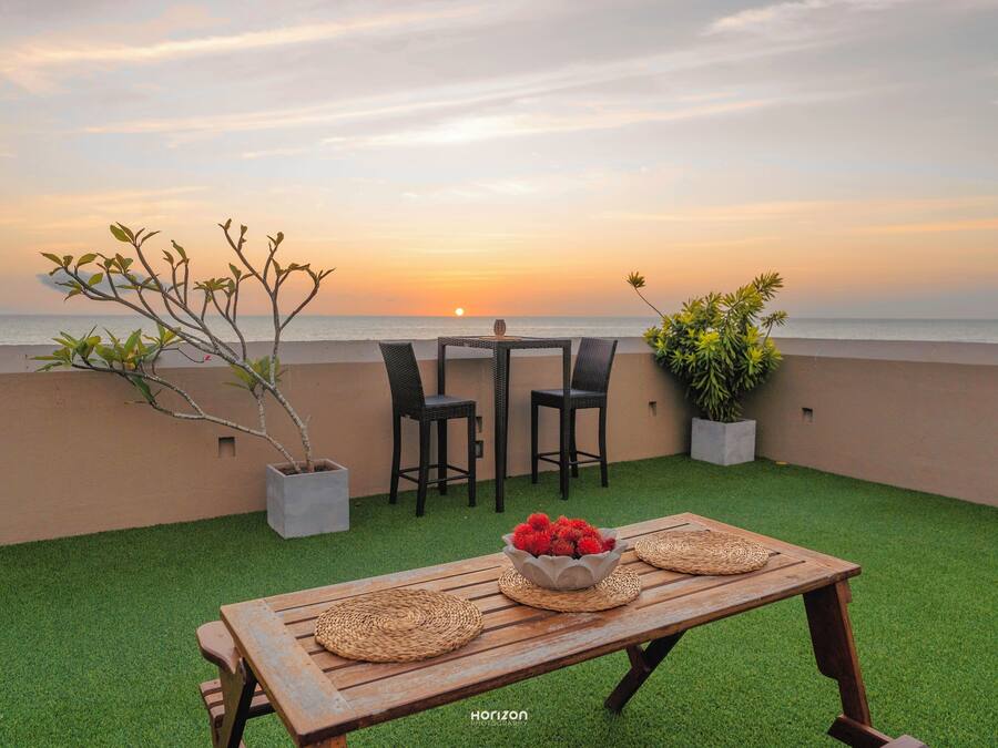 Um terraço com uma mesa de madeira, duas cadeiras e vista para o pôr do sol sobre o oceano.