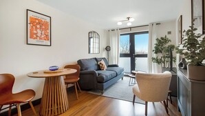 Uma sala de estar moderna com sofá, cadeiras, uma mesa de madeira redonda e uma janela grande com cortinas.
