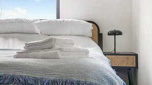 Cama bem arrumada com lençóis brancos e uma toalha dobrada, mesa de cabeceira com abajur, e janela com vista para o céu.
