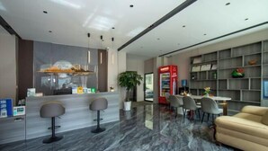 Um lobby moderno de hotel com balcão de recepção, banquetas de bar, área de refeições com mesa redonda e cadeiras, sofá e estante de livros.