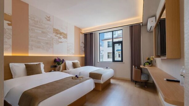 Quarto de hotel com duas camas, uma escrivaninha, uma cadeira, janela com cortinas e televisão na parede.