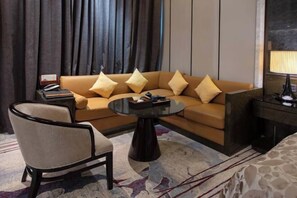 Quarto de hotel moderno com um sofá bege, uma mesa de centro redonda, uma mesinha de cabeceira e uma cama com colcha estampada.