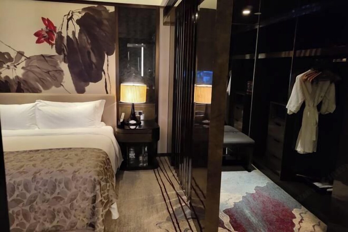 Quarto de hotel com cama, mesa de cabeceira com abajur, espelho e cabideiro.