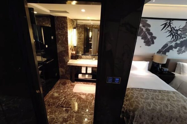 Quarto de hotel com chão de mármore, cama com cobertura estampada, mesa de cabeceira com abajur, banheiro com espelho e pia, e televisão.