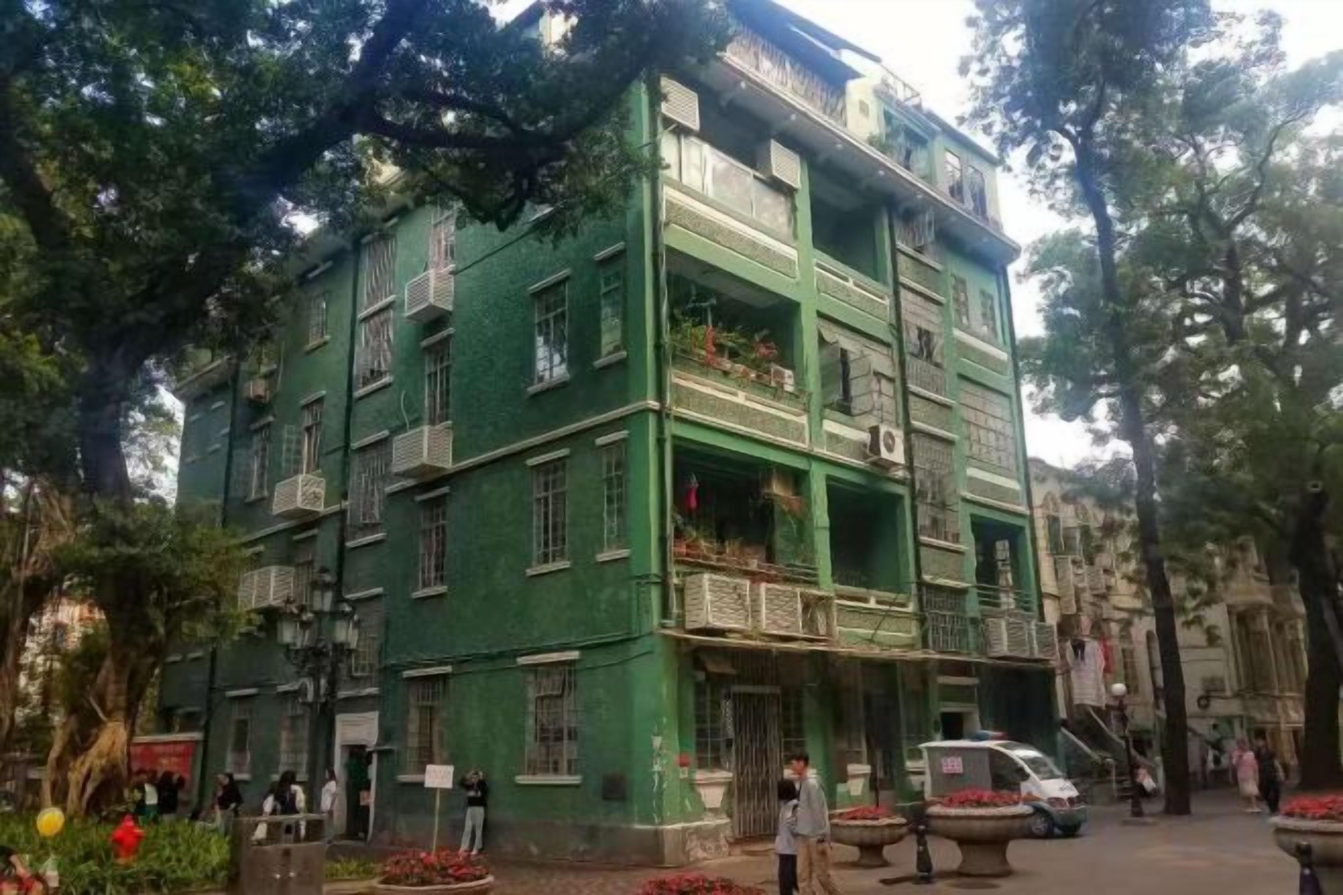 Um edifício com várias andares e varandas, com uma rua à frente e pessoas passando.