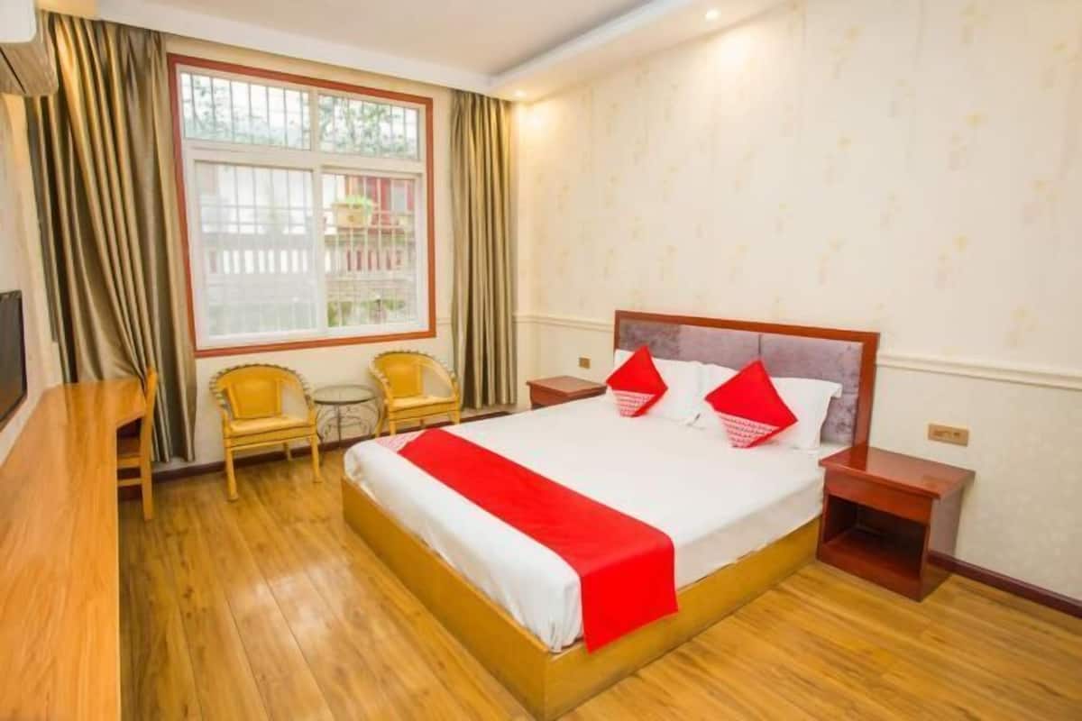 Quarto de hotel com uma cama grande, uma mesinha, uma cadeira, uma janela com cortinas e uma mesa de cabeceira.