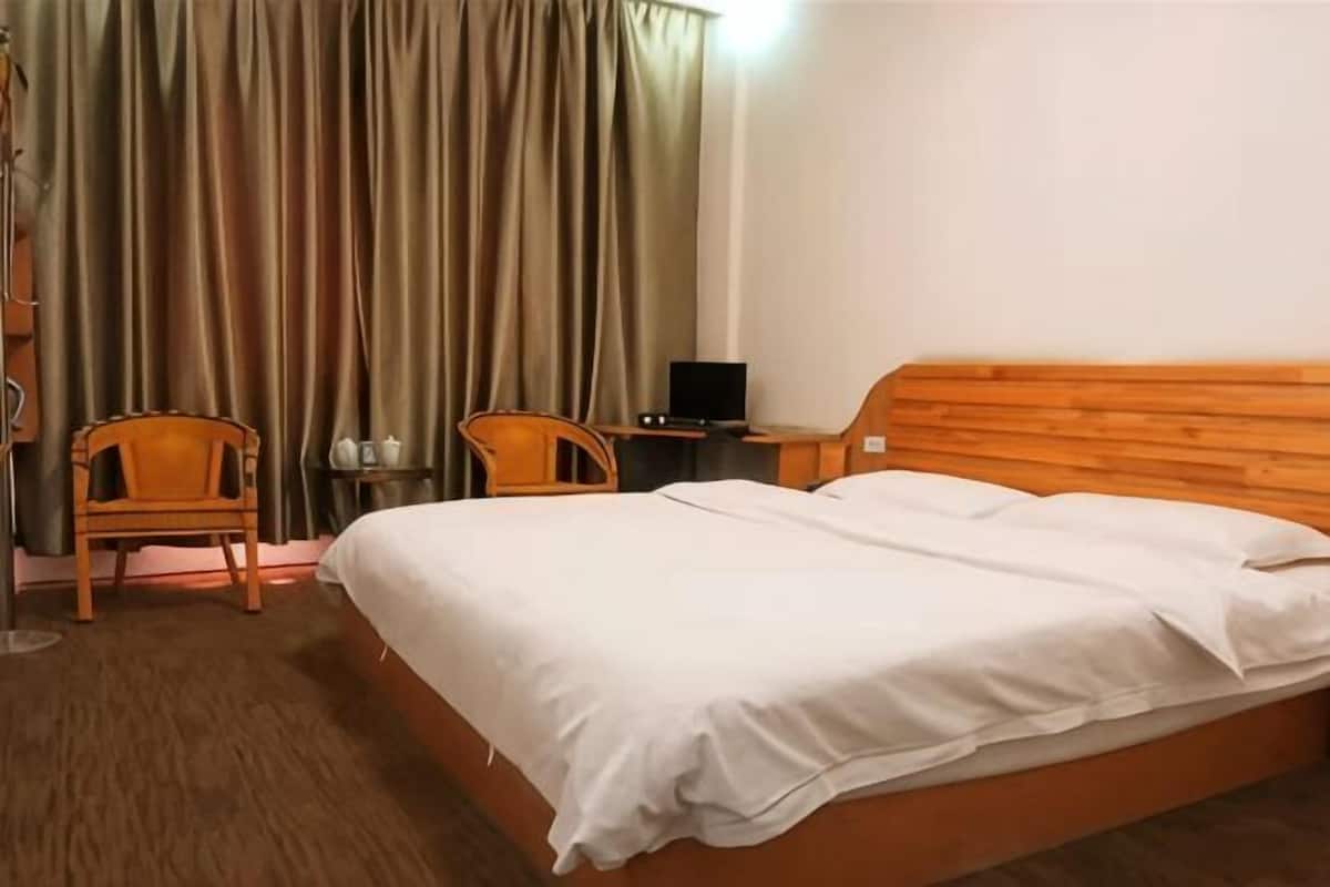 Quarto de hotel com uma cama de madeira, duas cadeiras, uma escrivaninha com um computador e uma cortina.