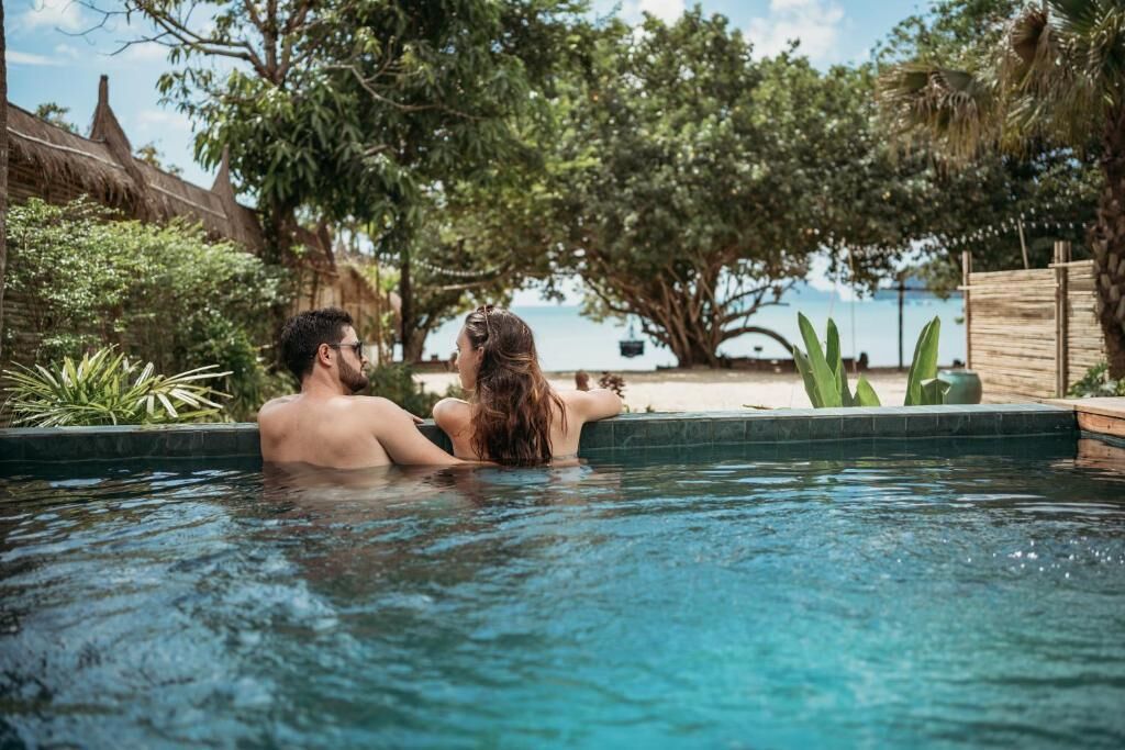 Um casal relaxando em uma piscina, com um cenário tropical, incluindo árvores e uma praia ao fundo.
