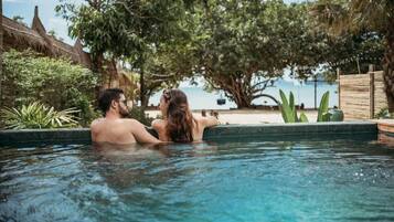 Um casal relaxando em uma piscina, com um cenário tropical, incluindo árvores e uma praia ao fundo.