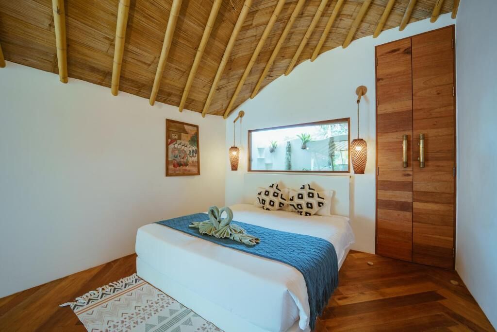 Quarto com teto de madeira, cama com manta azul, almofadas estampadas, luminária fixada na parede e um guarda-roupa.