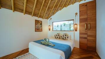 Quarto com teto de madeira, cama com manta azul, almofadas estampadas, luminária fixada na parede e um guarda-roupa.