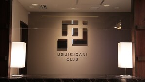 Uma área de recepção com design moderno, exibindo um logotipo e o nome 'UGUISUDANI CLUB' na parede. Há duas luminárias de pé de cada lado do balcão de recepção.