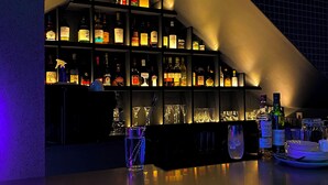 Uma área de bar com prateleiras cheias de garrafas de bebidas, copos e balcão.