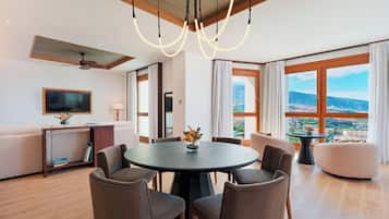 Uma zona de refeições moderna com uma mesa redonda, seis cadeiras, um candelabro, uma televisão na parede e grandes janelas com vista para a cidade.