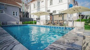Uma piscina com borda de pedra, dois guarda-sóis e um edifício branco ao fundo.