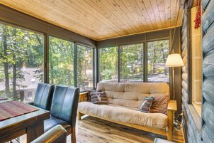 Interior acolhedor de uma cabana com sofá, cadeiras e grandes janelas com vista para a floresta ao redor.