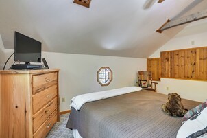Quarto com cama, cómoda de madeira, televisão sobre a cómoda, janela e vigamento de madeira no teto.