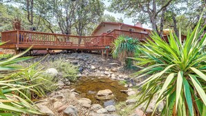 Uma ponte de madeira sobre um pequeno riacho, cercada por pedras e vegetação, conduzindo a uma casa com telhado vermelho.